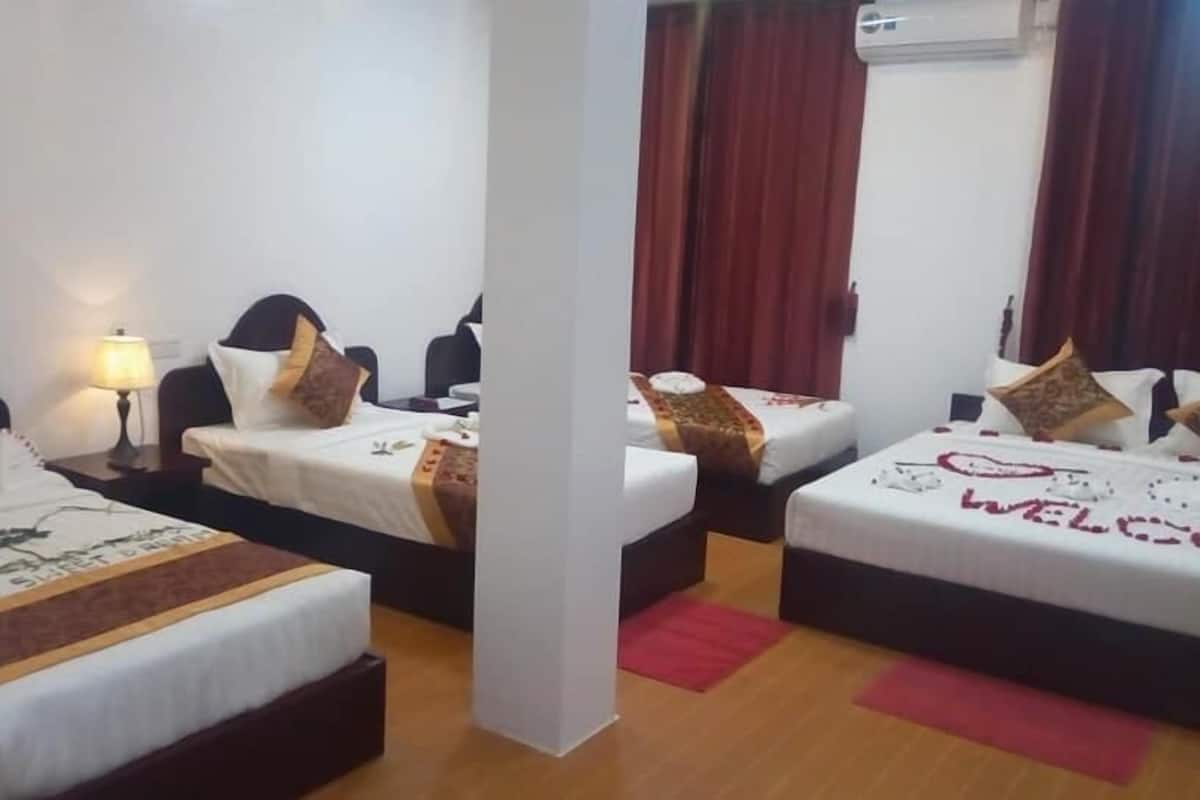 Quarto de hotel com três camas, cada uma com colcha branca e almofadas decorativas. Há uma mesa de cabeceira com luminária, um tapete vermelho no chão e um aparelho de ar condicionado na parede.