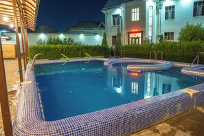 Uma piscina com borda azulejada e uma banheira de hidromassagem, rodeada por um deck de madeira e um edifício com um letreiro iluminado ao fundo.
