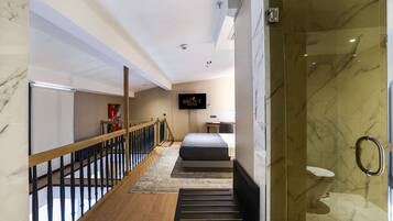 Quarto de hotel com uma cama, escrivaninha, televisão e banheiro com chuveiro de mármore.