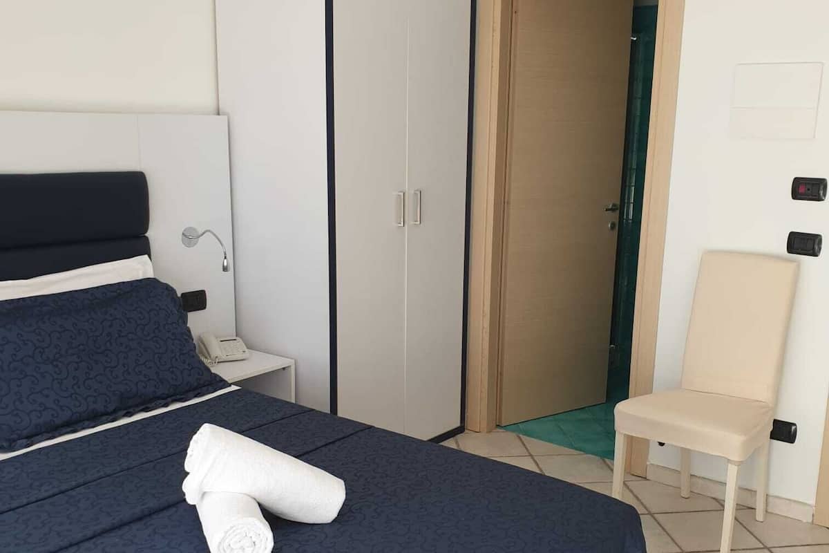Quarto de hotel com cama, guarda-roupa, cadeira e pequena mesa com telefone.