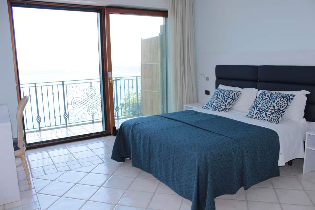 Quarto com cama, mesa de cabeceira, janela com vista e varanda.