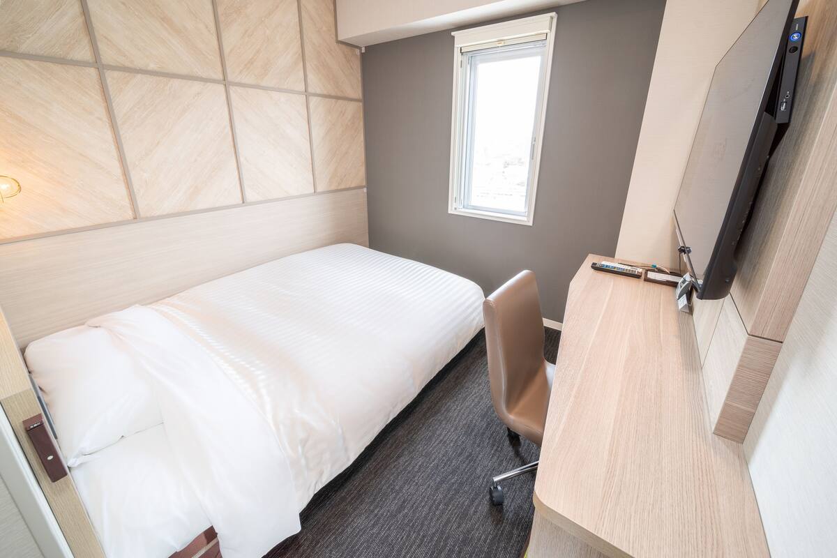 Quarto de hotel com cama, escrivaninha, cadeira, televisão e janela.