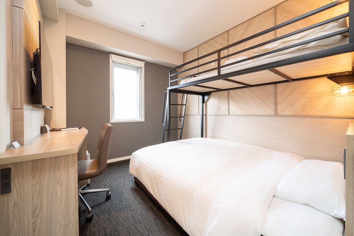 Quarto de hotel moderno com beliche, escrivaninha com cadeira, pequena janela e televisão de tela plana na parede.