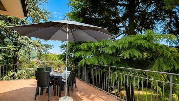 Uma área de refeições ao ar livre com mesa, cadeiras, um guarda-sol grande e um corrimão de metal.