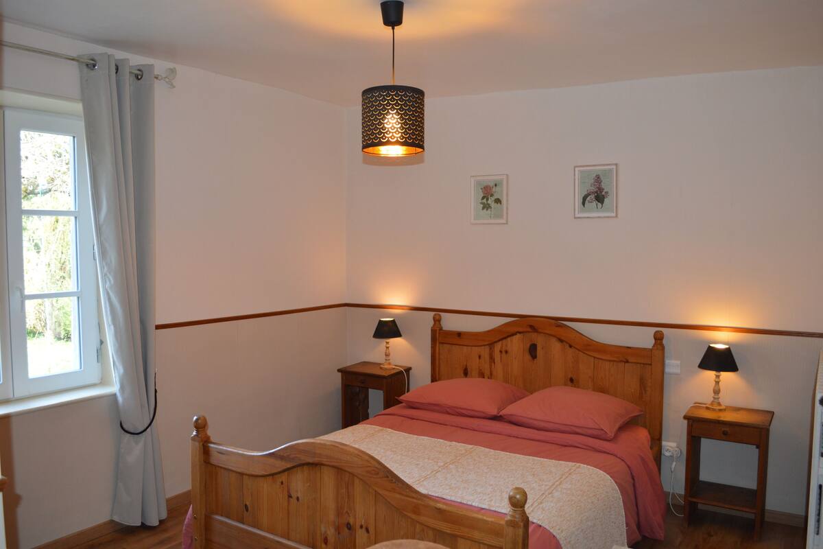 Quarto com cama de madeira, duas mesas de cabeceira com luminárias, uma janela com cortinas e dois quadros na parede.