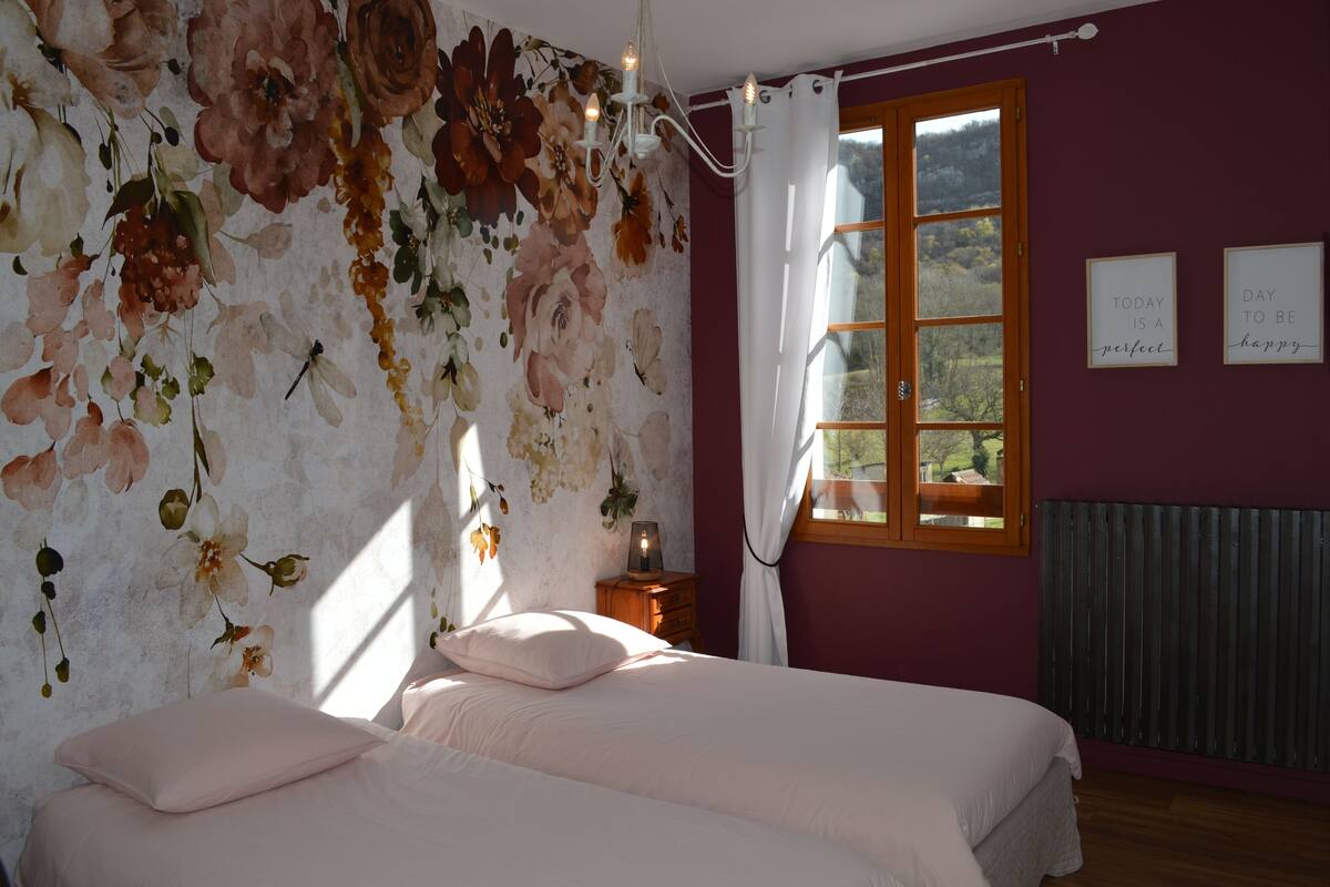 Quarto com papel de parede floral, cama com roupa de cama branca, mesa de cabeceira com abajur e janela com vista para o exterior.