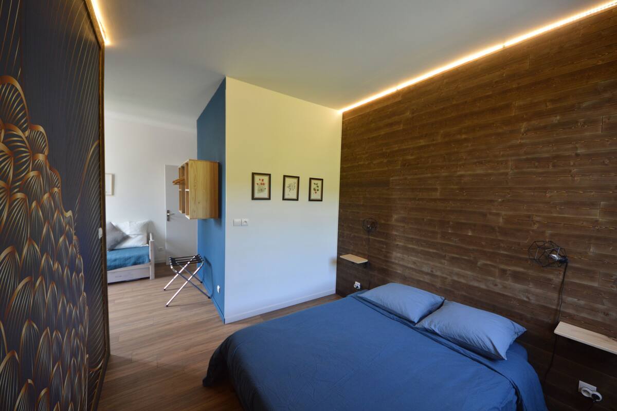 Um quarto moderno com cabeceira de madeira, cama com roupa de cama azul, um pequeno espaço de estar com uma cadeira e uma mesa, e uma prateleira fixada na parede.
