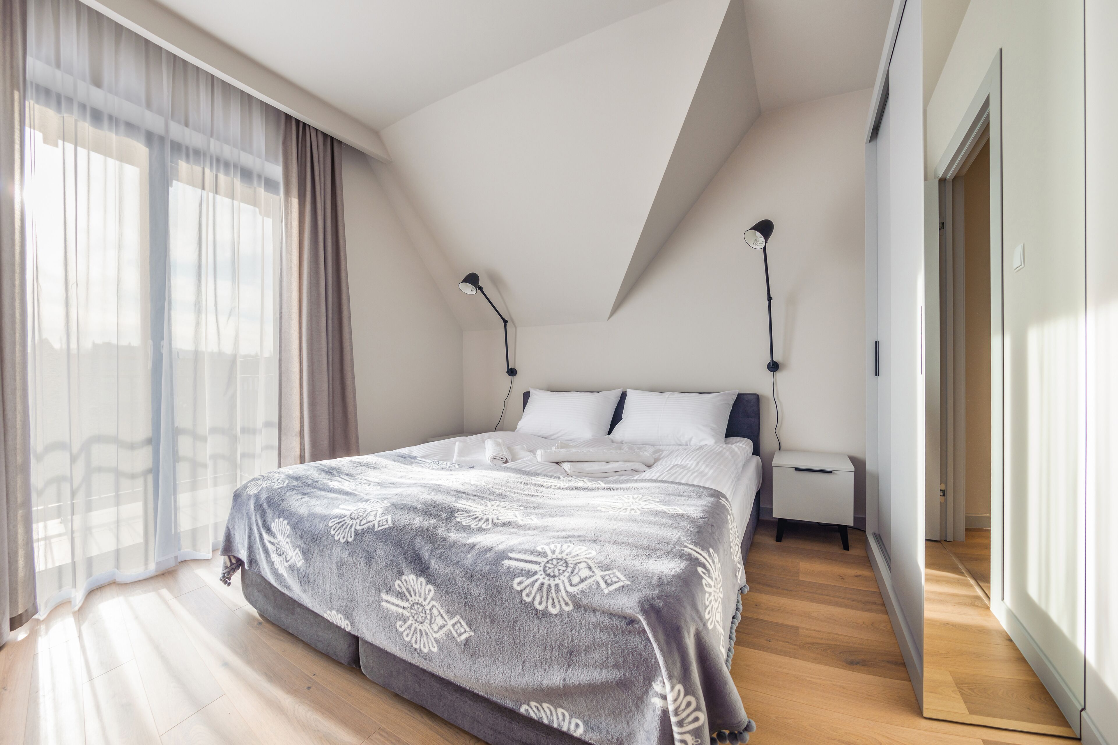 Um quarto com cama, mesas de cabeceira, um criado-mudo, luminária e uma janela com cortinas leves.
