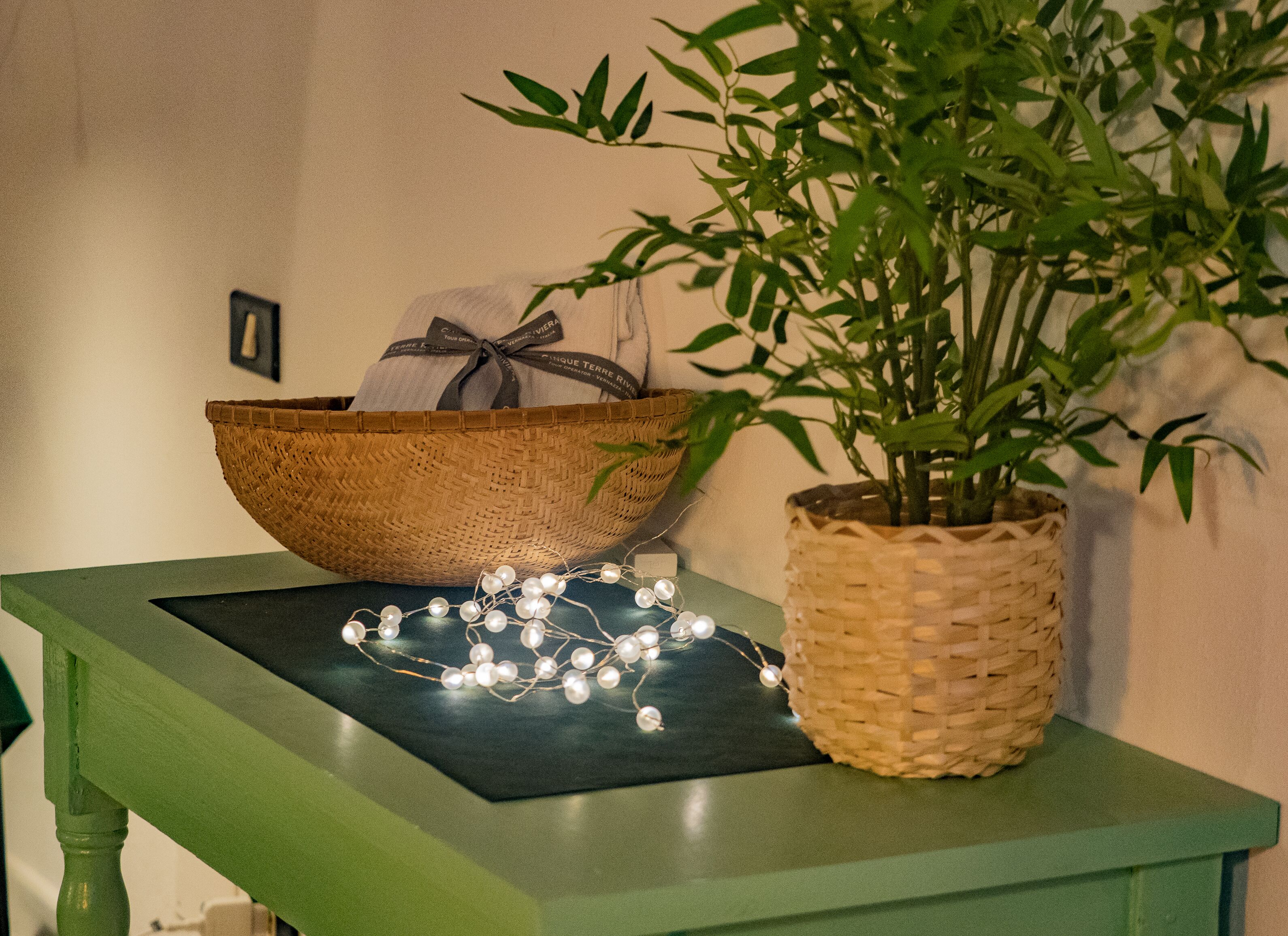 Uma mesa verde com uma cesta de palha, uma planta em vaso e um colar de pérolas.