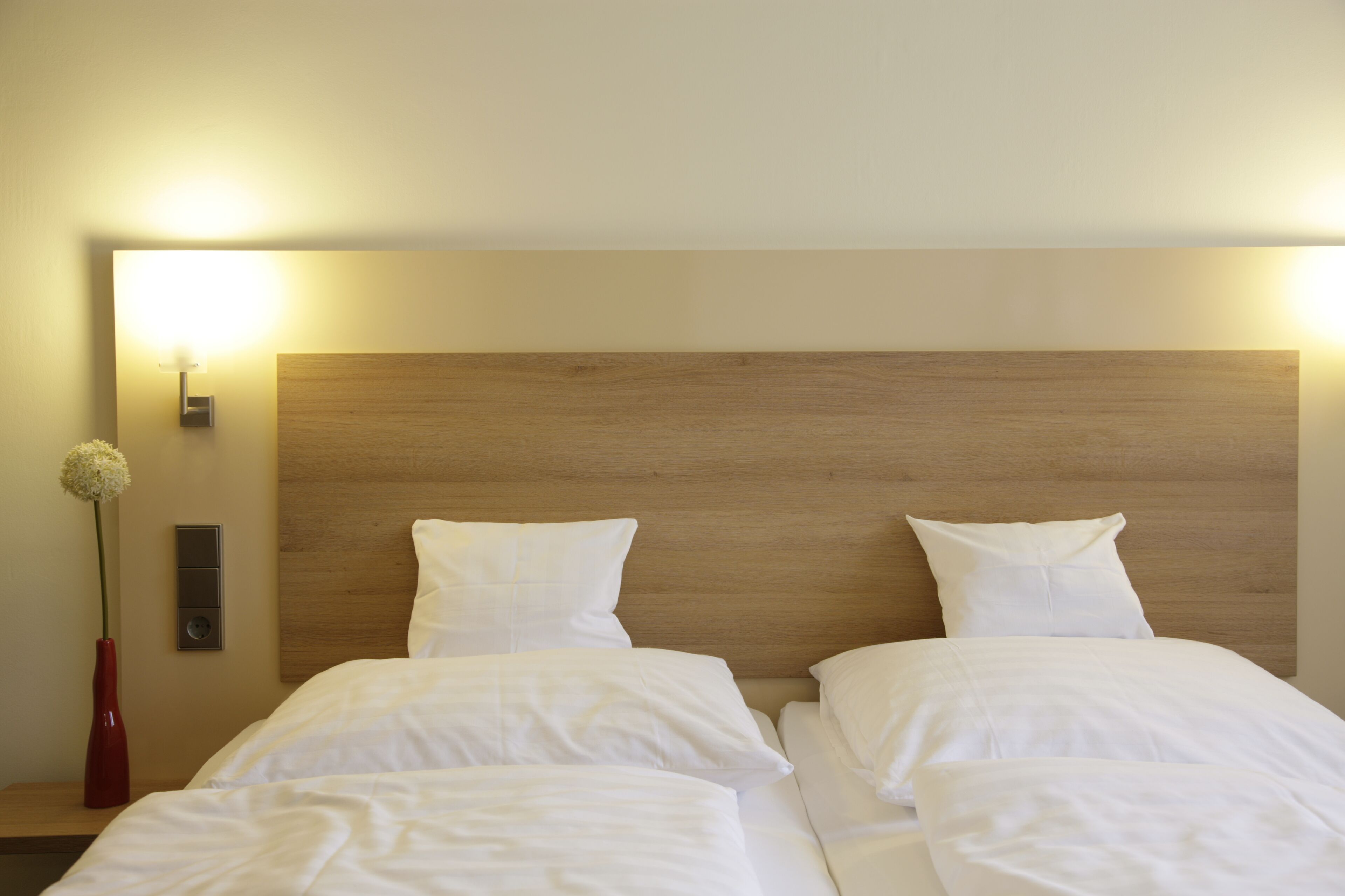 Quarto de hotel com cabeceira de madeira, duas camas com roupa de cama branca, um vaso vermelho com flores e um candeeiro fixo na parede.