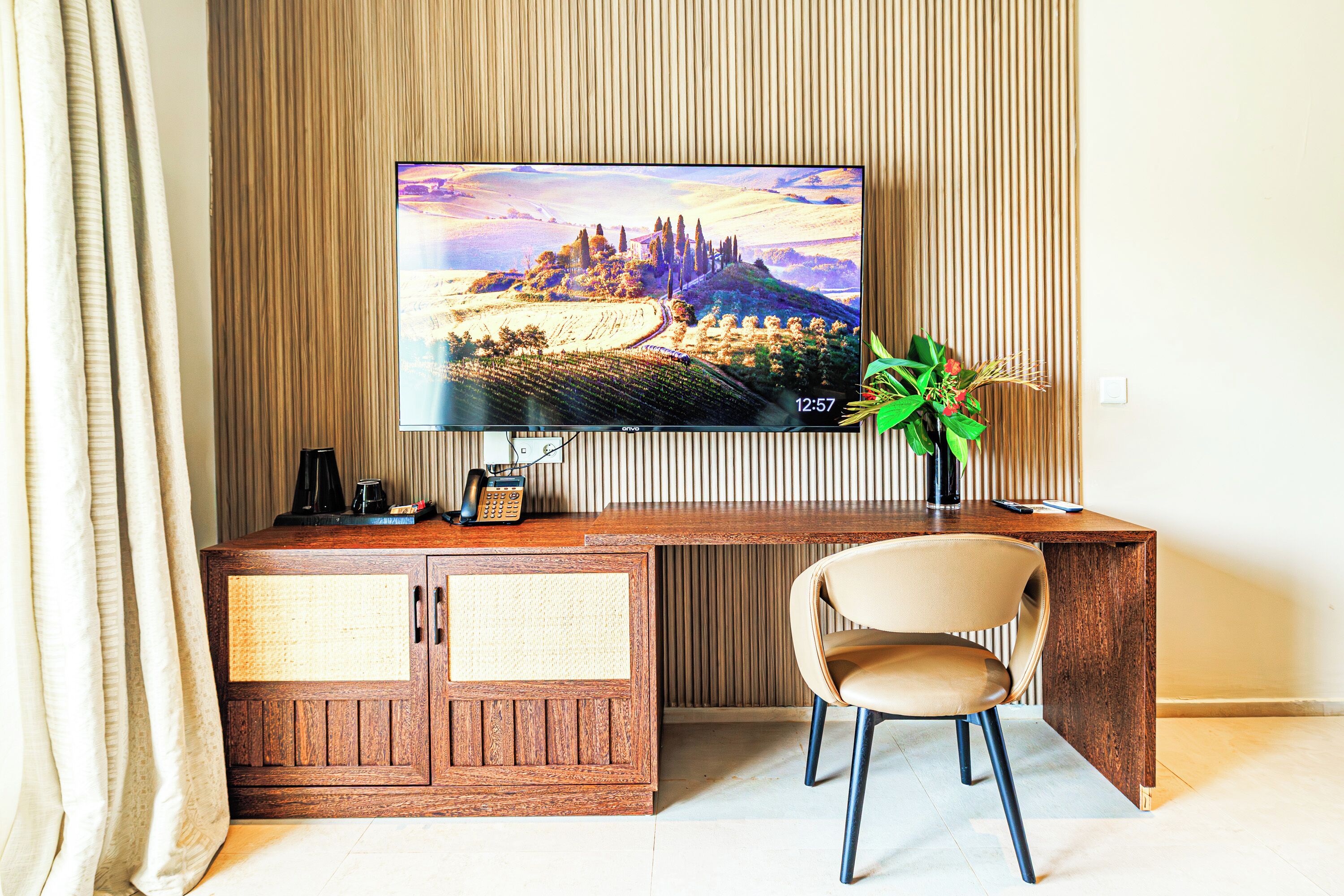 Quarto de hotel com uma secretária de madeira, uma cadeira, uma televisão presa à parede e uma planta na secretária.