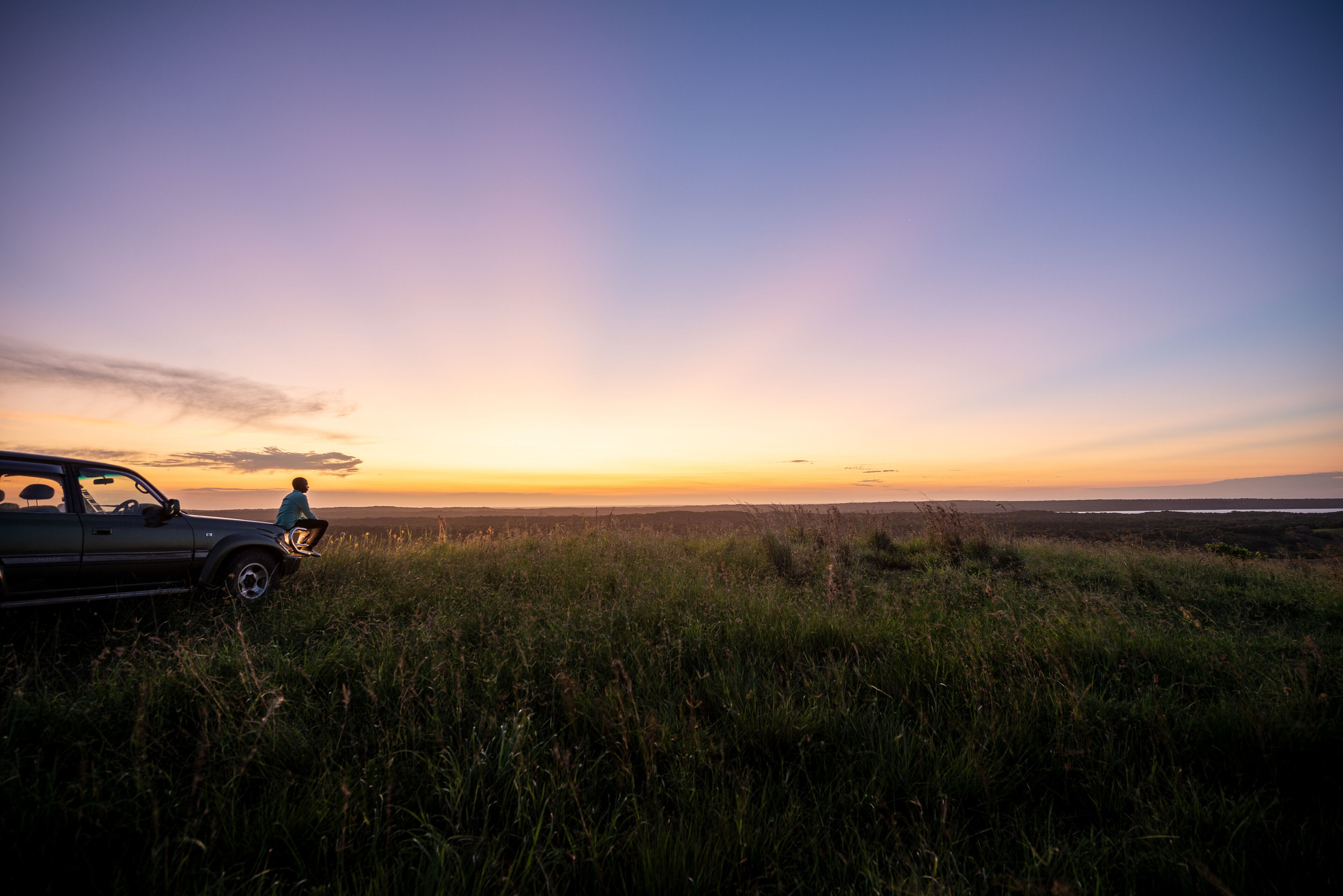 Um carro estacionado em um campo gramado, com uma pessoa ao lado observando o pôr do sol.