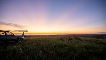 Um carro estacionado em um campo gramado, com uma pessoa ao lado observando o pôr do sol.
