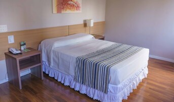 Quarto de hotel com uma cama, uma mesa de cabeceira e um telefone, e uma luminária fixada na parede.