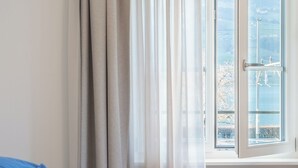 Um quarto com cama, janela com vista para um corpo de água e montanhas, e cortina.