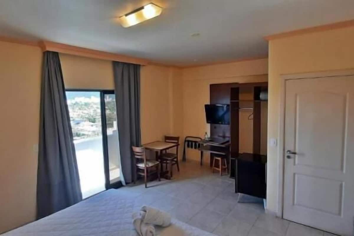 Quarto de hotel com cama, escrivaninha, cadeira, televisão e porta.