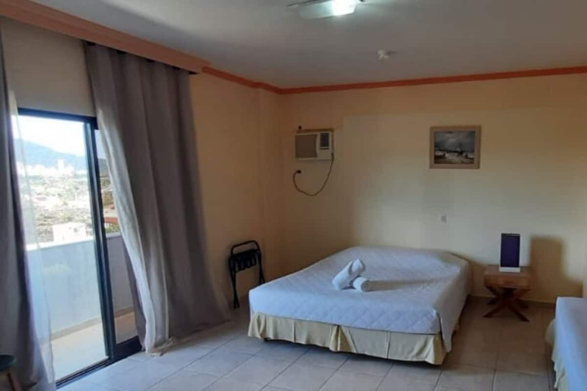 Quarto de hotel com cama, uma cadeira, uma mesinha, guarda-roupa e janela com cortinas.
