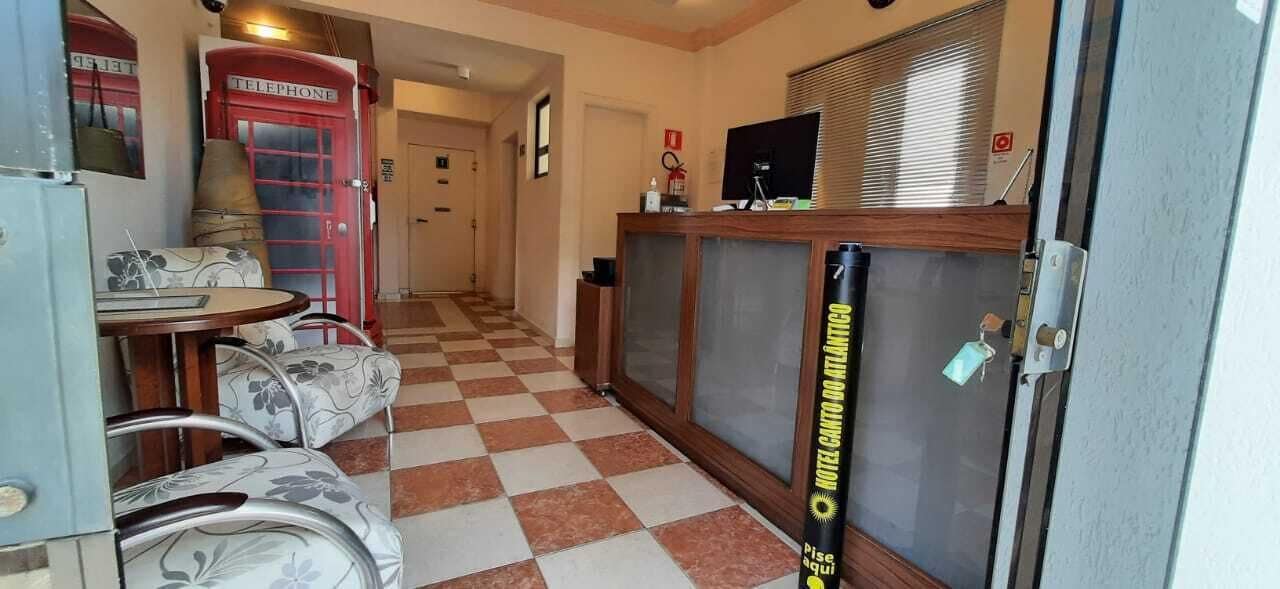 Um saguão de hotel com piso xadrez, uma cabine telefônica vermelha, um armário de madeira com uma placa e uma pequena mesa com cadeiras.