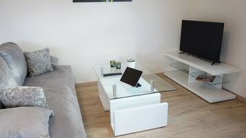 Uma sala de estar moderna com um sofá cinzento, uma mesa de centro de vidro com um laptop, uma TV em um suporte branco e uma pequena planta.