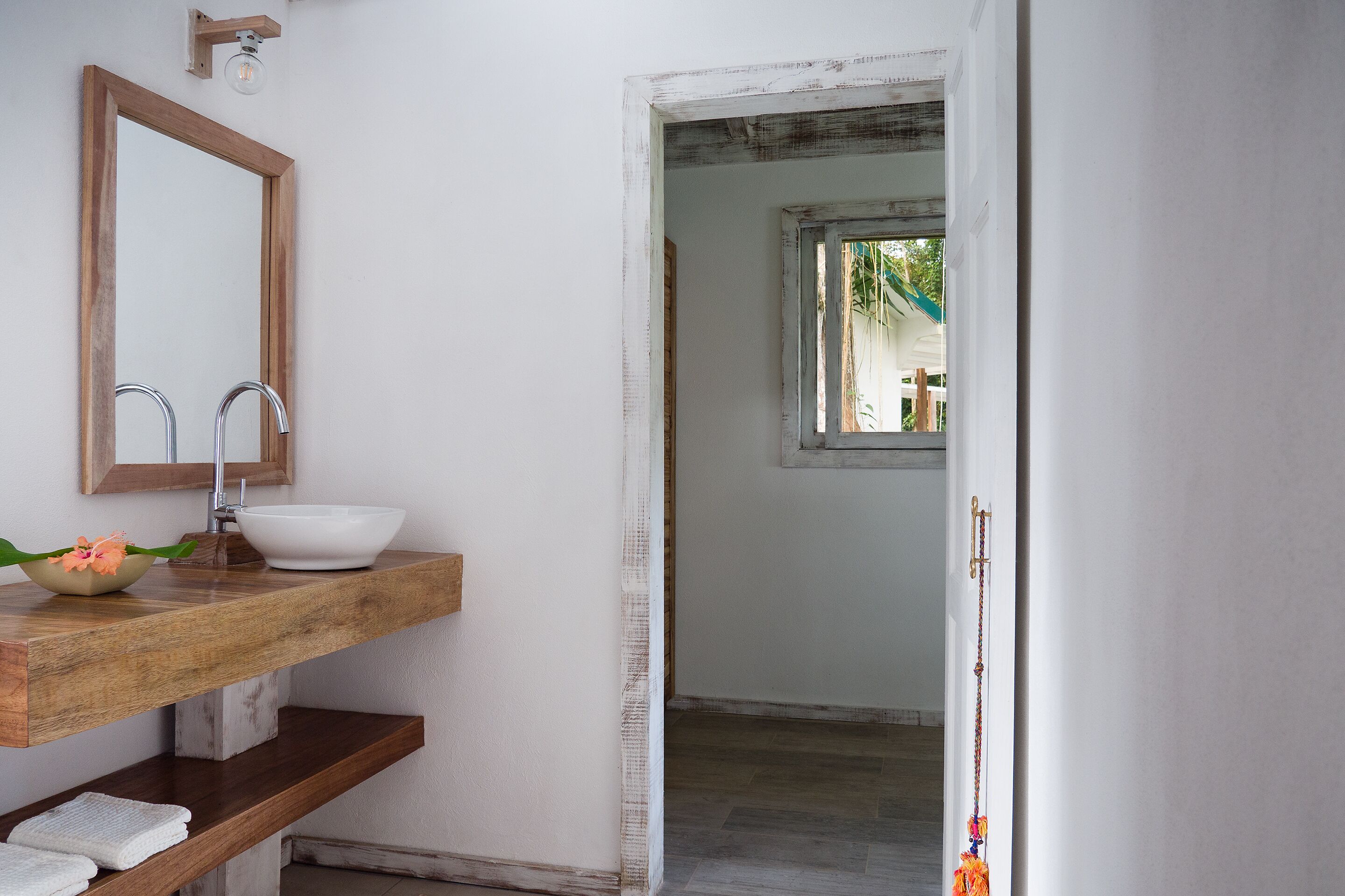 Casa de banho com toucador de madeira, lavatório branco e espelho. Há uma janela com vista para o verde lá fora.