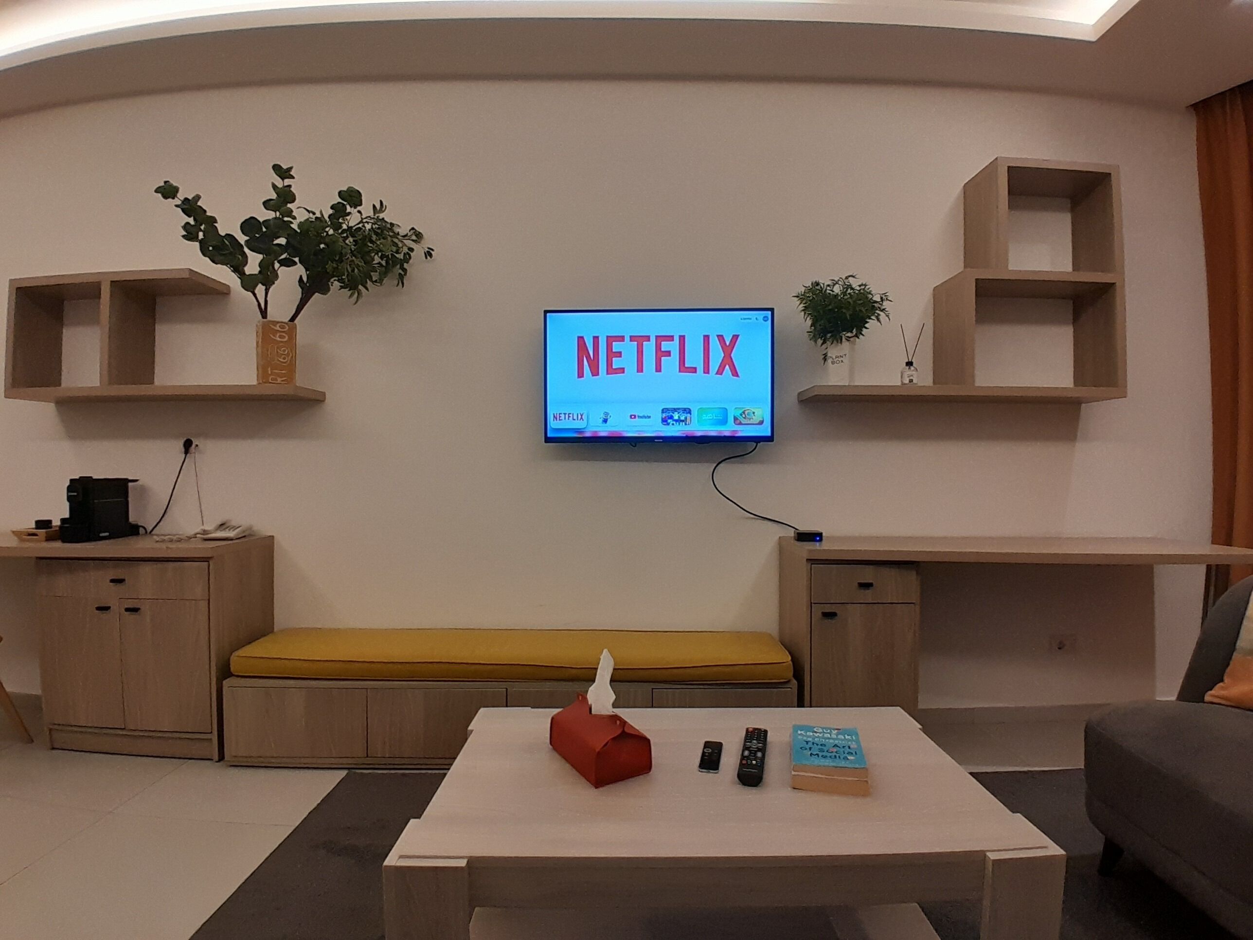 Uma sala de estar moderna com uma TV de tela plana exibindo o logotipo da Netflix, um banco de madeira, uma mesa de centro com controles remotos e um livro, e estantes embutidas com plantas.