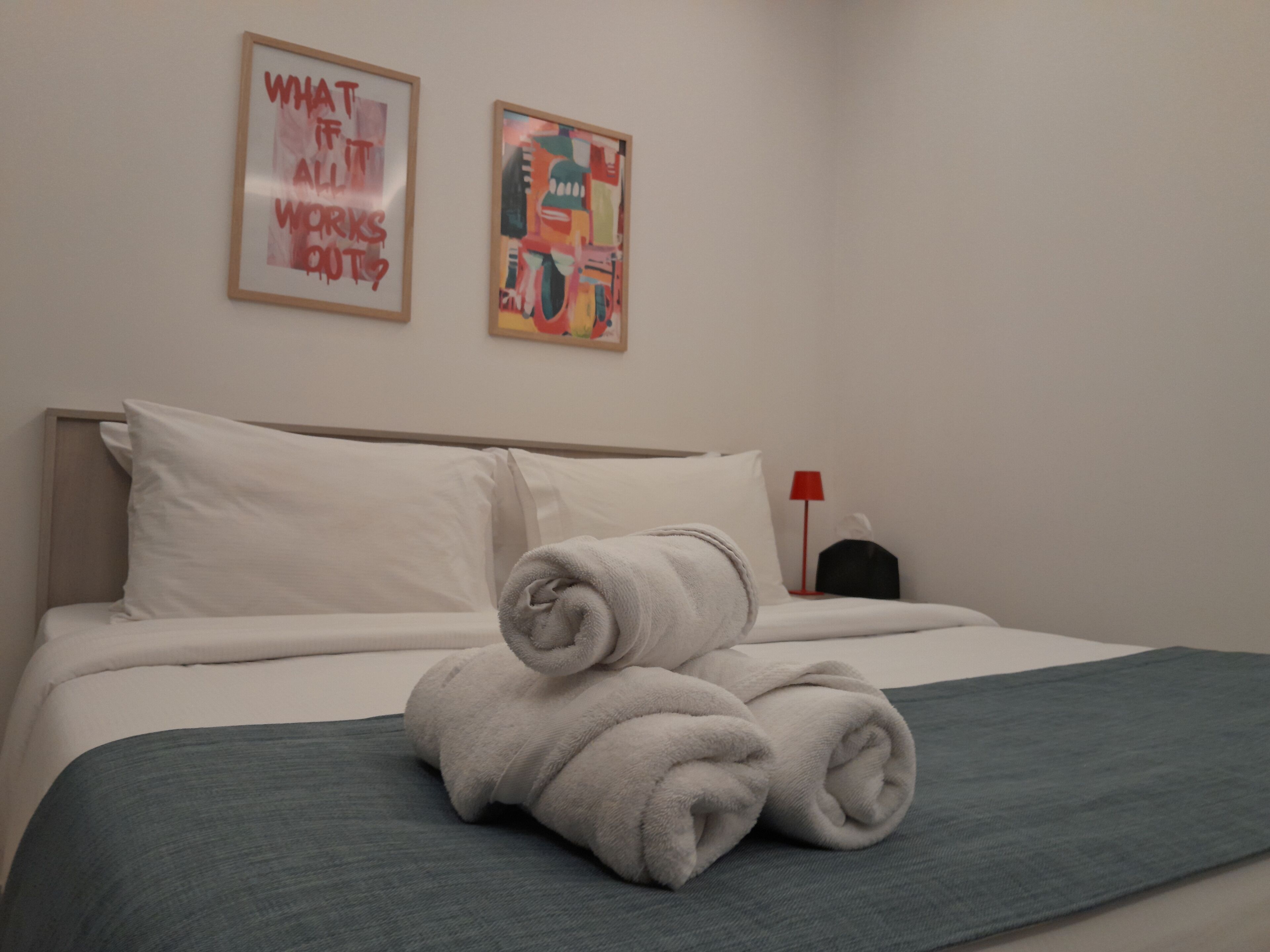 Uma cama bem arrumada com lençóis brancos e um cobertor azul-turquesa, duas obras de arte emolduradas na parede e uma luminária vermelha na mesa de cabeceira.
