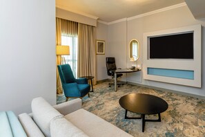 Quarto de hotel com sofá, poltrona, mesa pequena, escrivaninha, televisão e janela com cortinas.