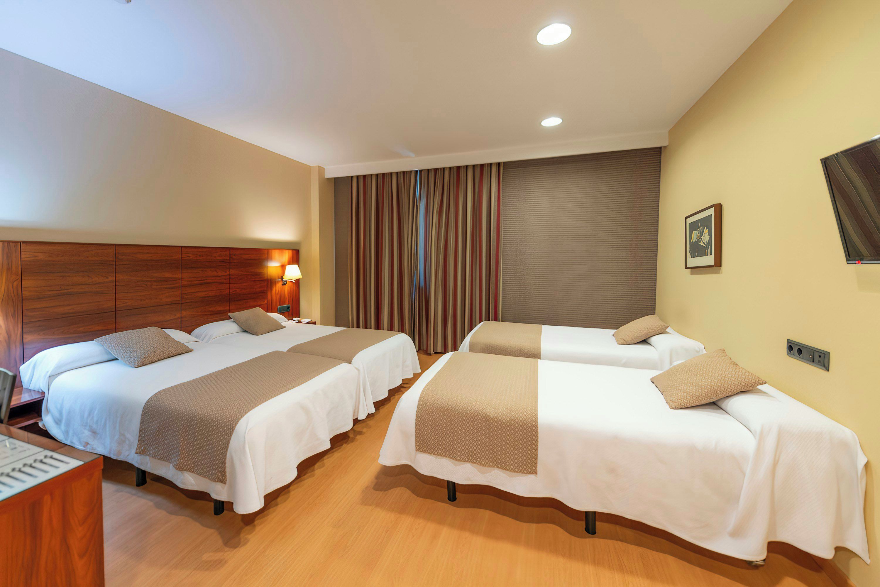 Quarto de hotel com duas camas de solteiro, cabeceira de madeira, escrivaninha com teclado e uma janela grande com cortinas.