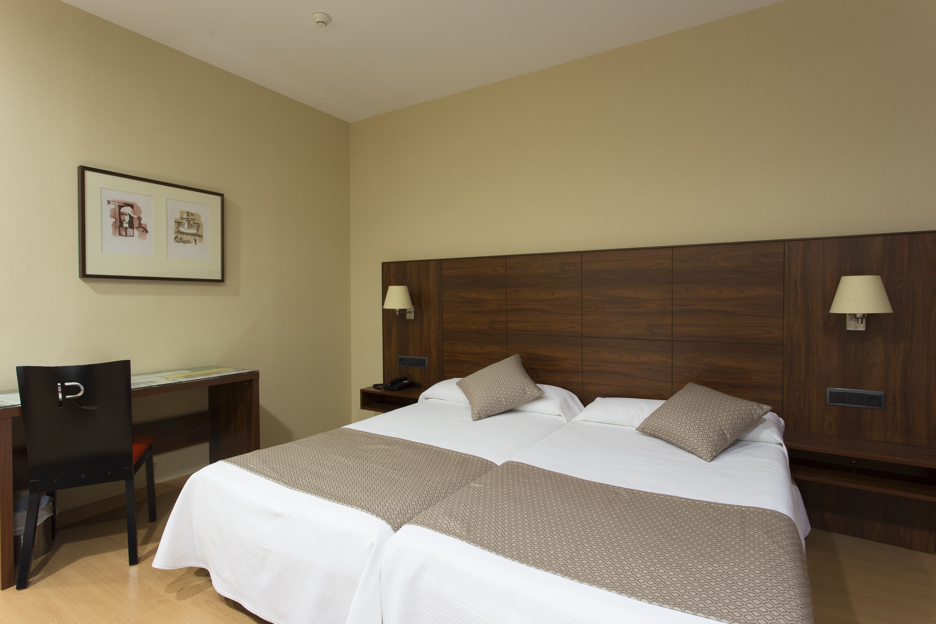 Quarto de hotel com uma cama grande, uma escrivaninha e duas cadeiras. Há duas mesas de cabeceira com abajures.