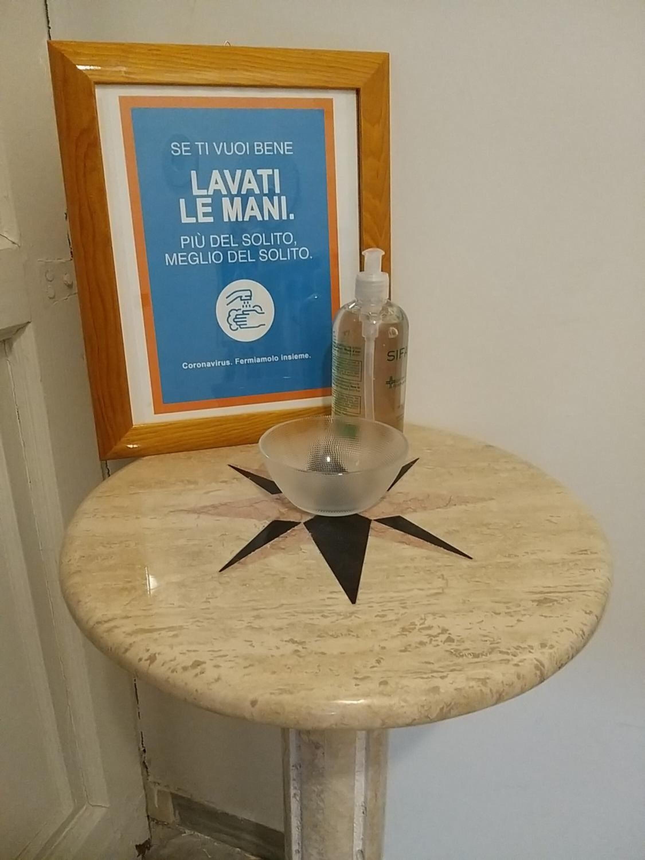 Uma pia de banheiro com um pôster emoldurado acima, um frasco de álcool em gel e um escorredor na pia.