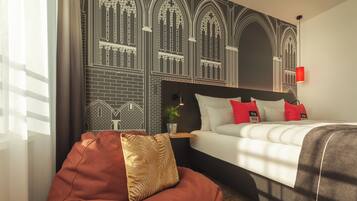 Quarto de hotel moderno com uma cama grande, uma poltrona de espuma vermelha e um mural na parede com projetos arquitetônicos.