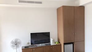Quarto com televisão sobre um móvel de madeira, um ventilador, um pequeno frigorífico e um cesto de lixo.