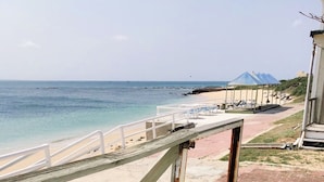 Um deck de madeira com vista para a praia, uma cadeira e um edifício com telhado azul ao fundo.