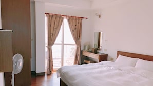 Quarto de hotel com cama, escrivaninha, cadeira, ventilador e janela com cortinas.