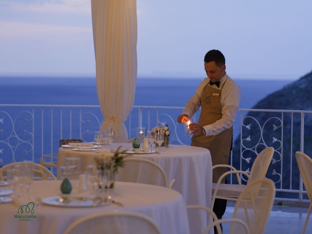Mesa posta para refeição com vista para o mar, um garçom atendendo e um corrimão de varanda.