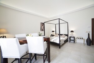 Quarto de hotel moderno com cama de dossel, mesa de jantar com cadeiras, pequena mesa de apoio, abajur e um vaso.
