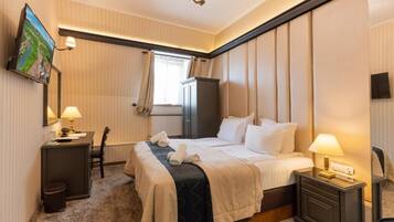 Quarto de hotel com cama, mesas de cabeceira, escrivaninha, cadeira, televisão e janela com cortinas.