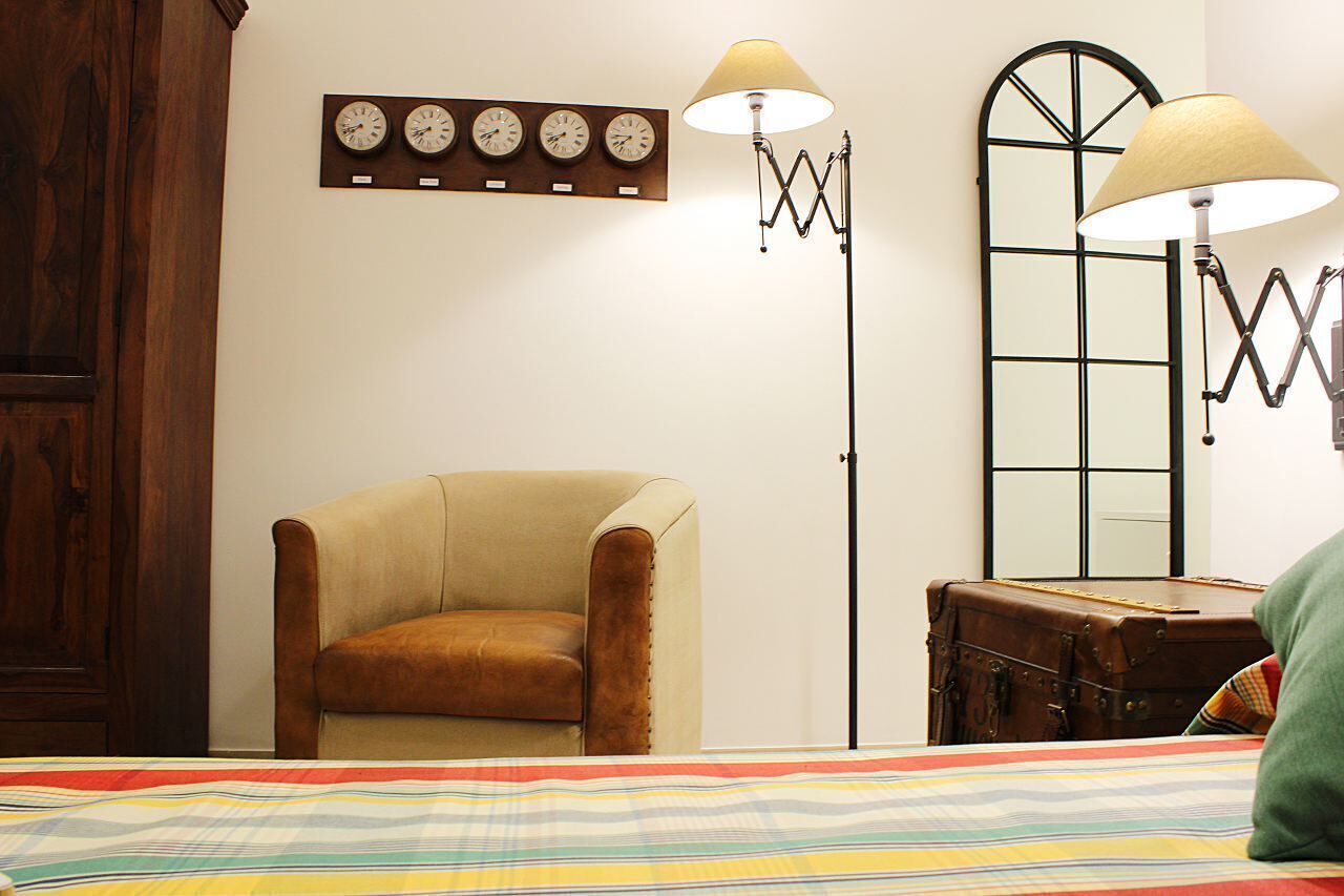 Quarto com cama, uma cadeira, relógio de parede, luminária de chão e mala.