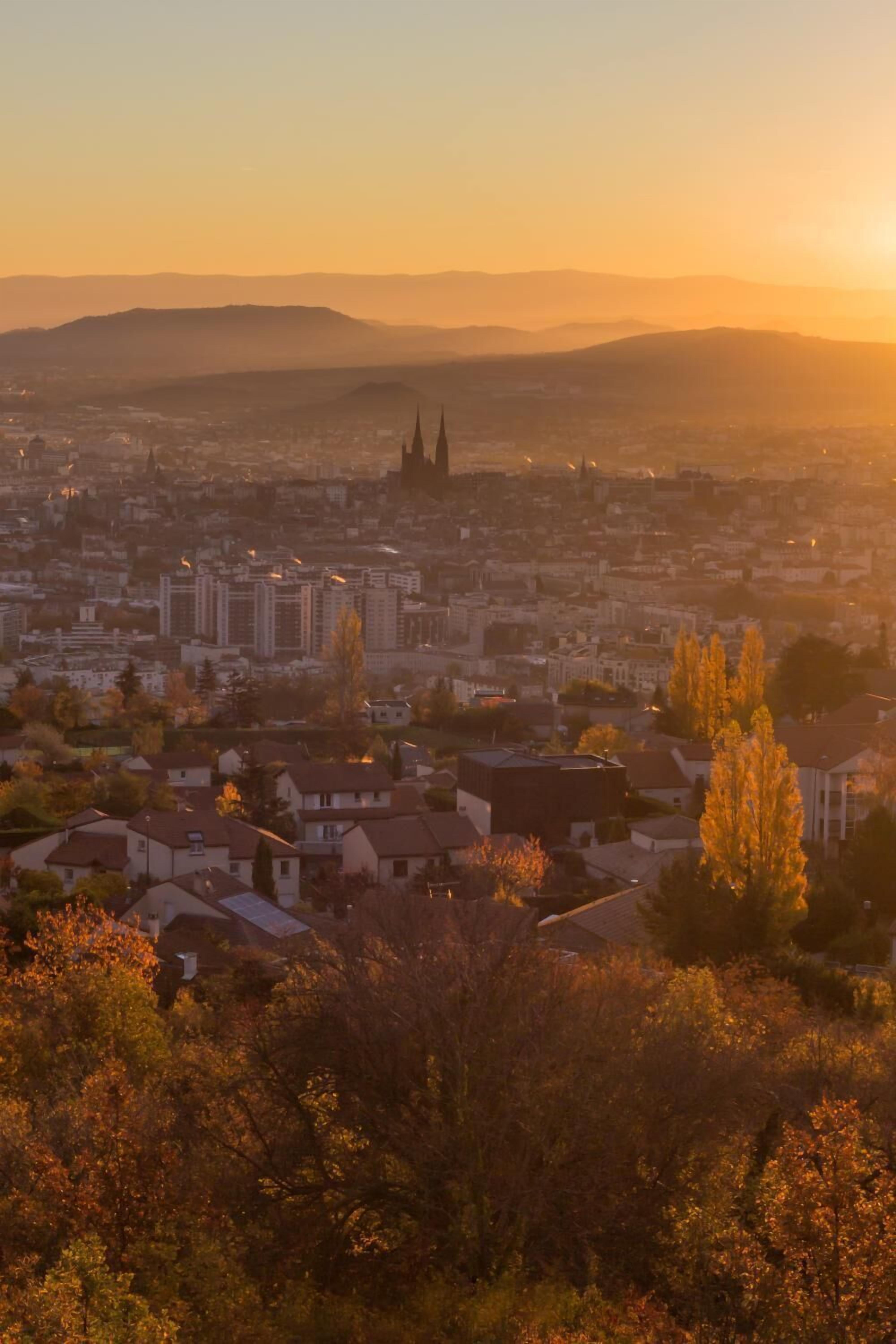 Paisagem urbana ao pôr do sol, com uma torre de igreja imponente, edifícios residenciais e um horizonte montanhoso.