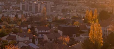 Paisagem urbana ao pôr do sol, com uma torre de igreja imponente, edifícios residenciais e um horizonte montanhoso.