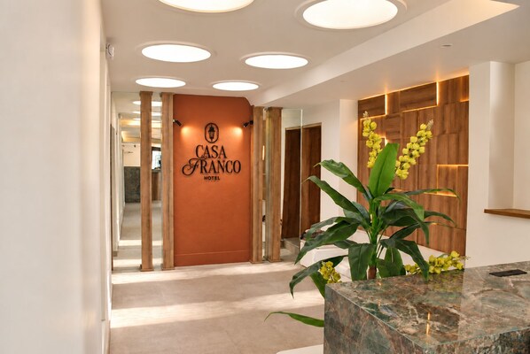 Uma área de recepção de hotel com uma placa indicando 'CASA FRANCO HOTEL' e um balcão de atendimento com uma planta.