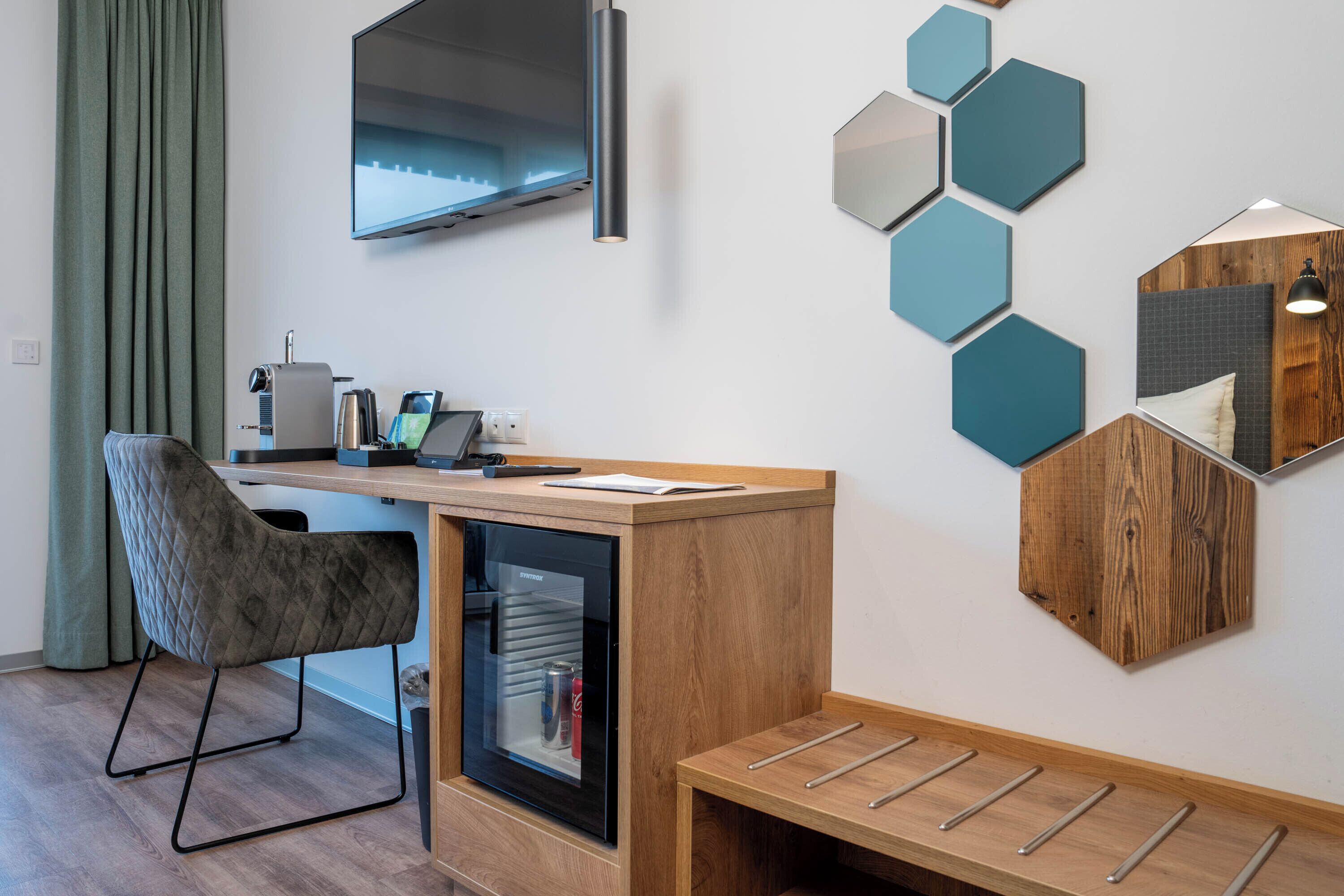 Quarto de hotel moderno com escrivaninha de madeira, cadeira confortável, mini-geladeira, TV de tela plana e um original ornamento hexagonal na parede.