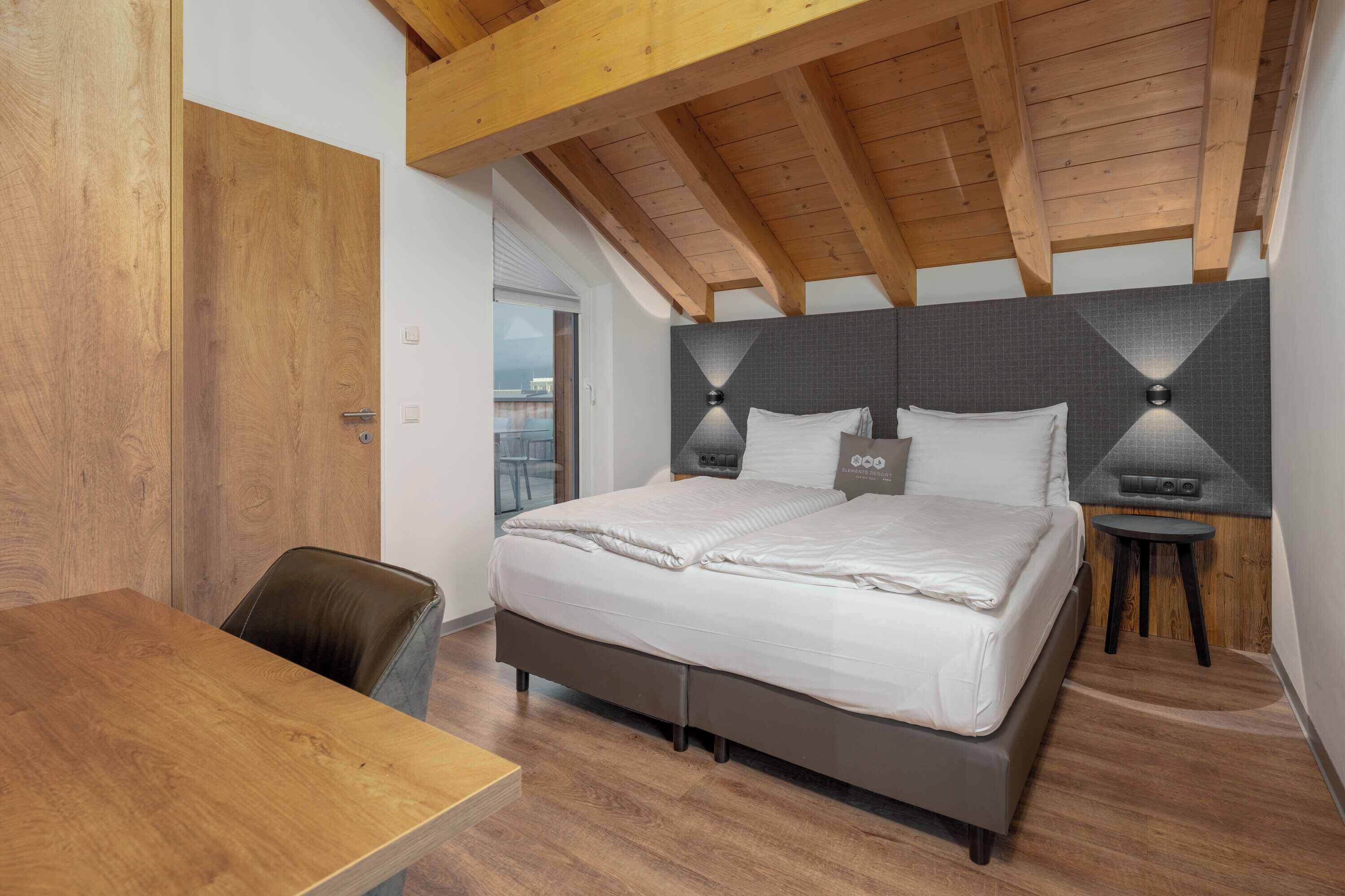 Quarto com teto de madeira, cama com cabeceira cinza, mesa de cabeceira e uma cadeira.