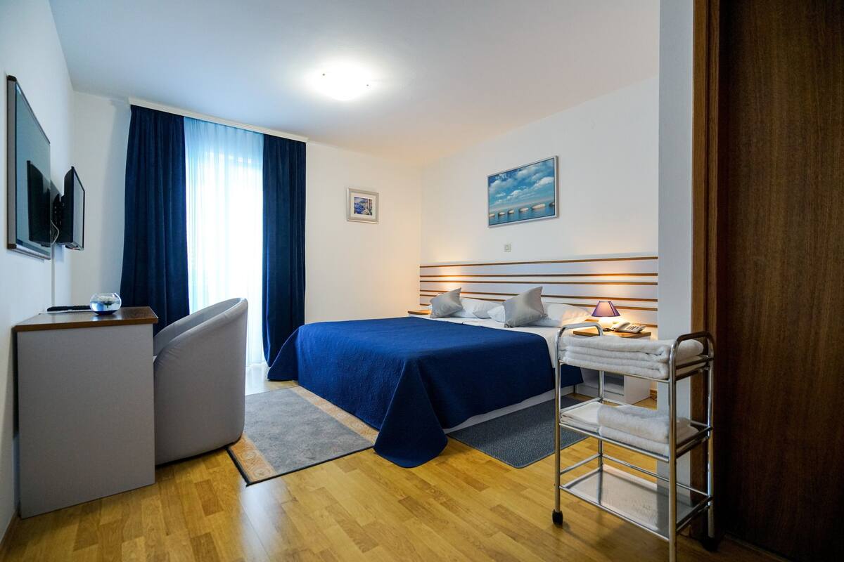 Quarto de hotel com cama, mesa de cabeceira, luminária e escrivaninha com cadeira.