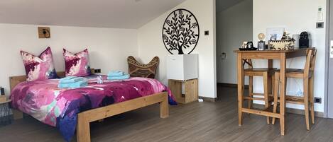 Quarto com cama, cabeceira de madeira, decoração de árvore fixada na parede, pequena mesa com chaleira e porta para outro cômodo.