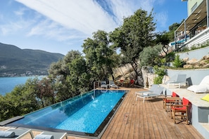 Uma piscina infinita retangular com um deck de madeira, espreguiçadeiras e uma mesa.