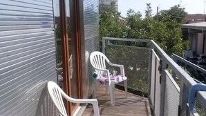 Uma varanda com duas cadeiras de plástico branco, corrimão de metal e vista para árvores e edifícios ao fundo.