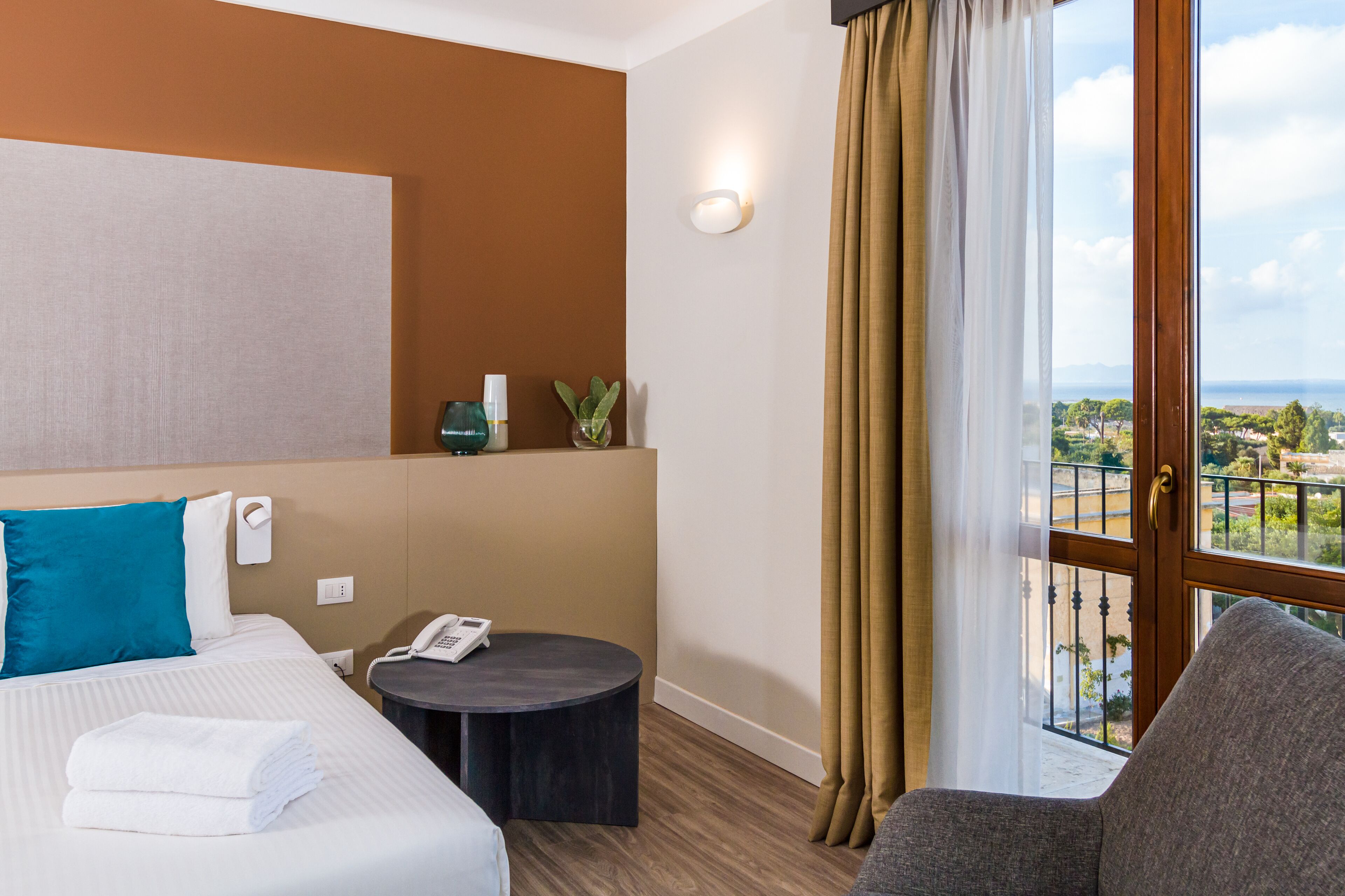 Quarto de hotel com uma cama, uma mesa redonda, uma cadeira, um telefone, uma luminária na parede e vista pela janela.