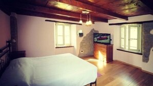 Quarto com cama, televisão, paredes de pedra, vigas de madeira e janelas amplas.