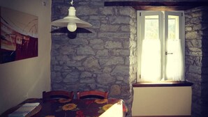 Uma área de refeições com parede de pedra, uma mesa com toalha de mesa de girassol, duas cadeiras, um lustre e uma janela com cortinas.
